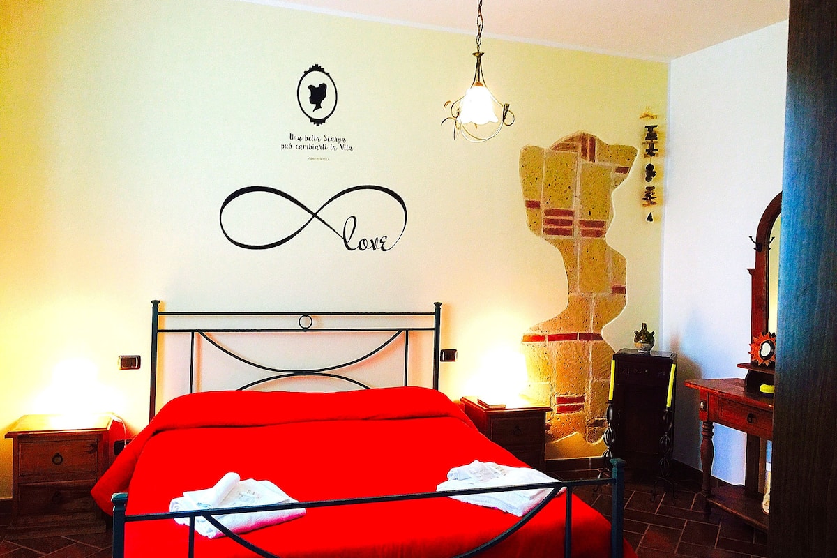 Quarto de hotel com cama vermelha, parede com símbolo de infinito em forma de coração e um objeto decorativo na parede.