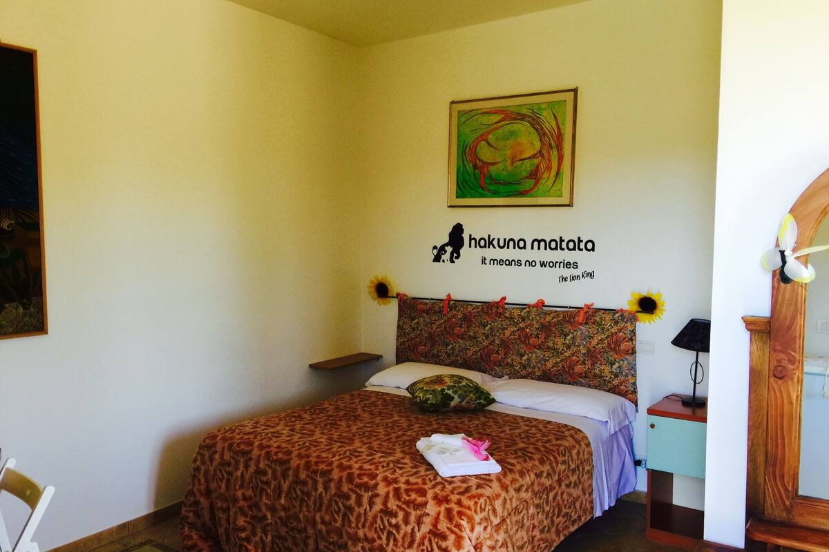 Quarto com cama, cabeceira com girassóis, uma obra de arte na parede e um letreiro com silhueta de leão e texto.