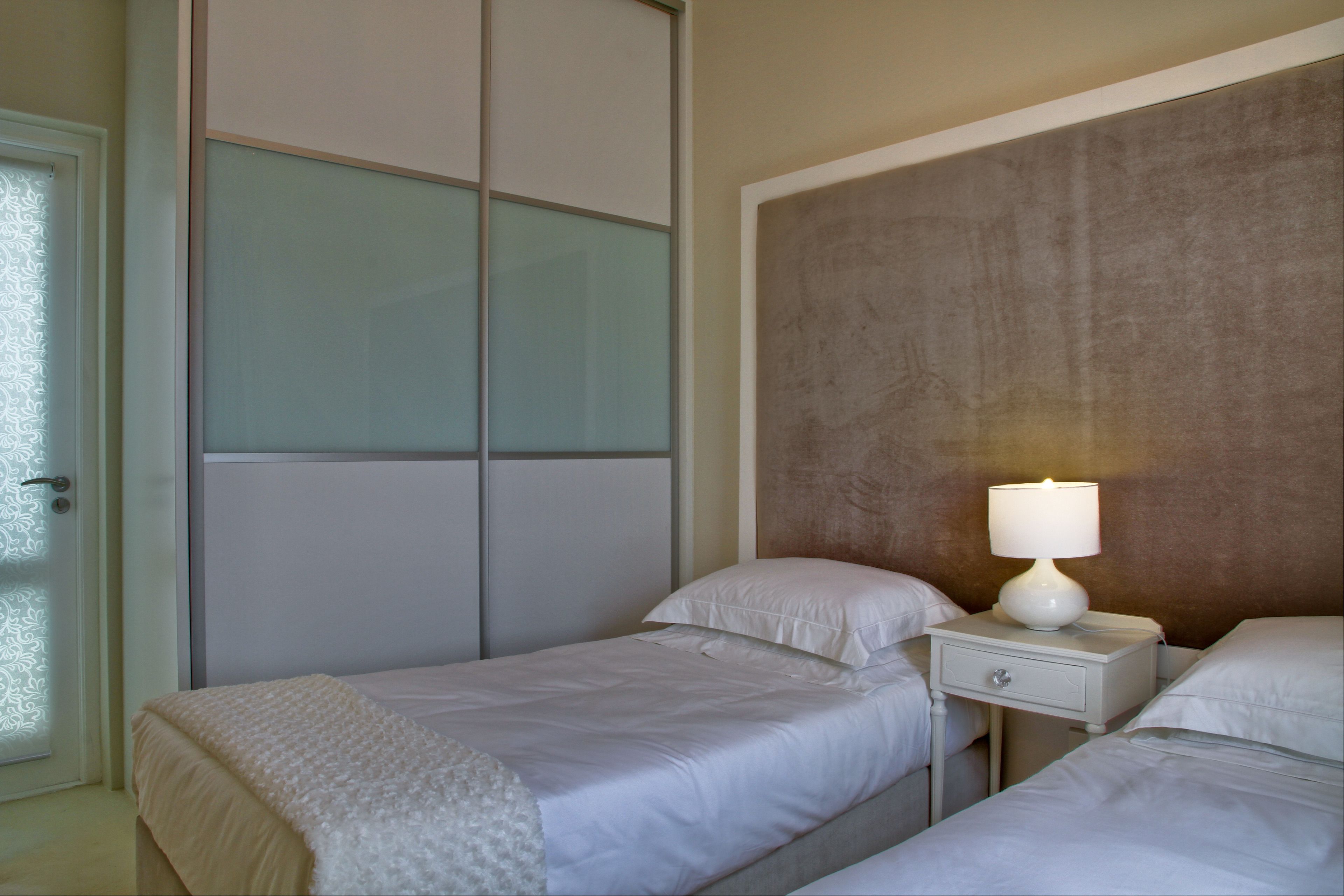 Quarto de hotel com duas camas de solteiro, mesa de cabeceira com luminária e guarda-roupa com portas de vidro jateado.