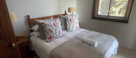 Quarto com cama, almofadas, mesa de cabeceira, abajur e janela com persianas.