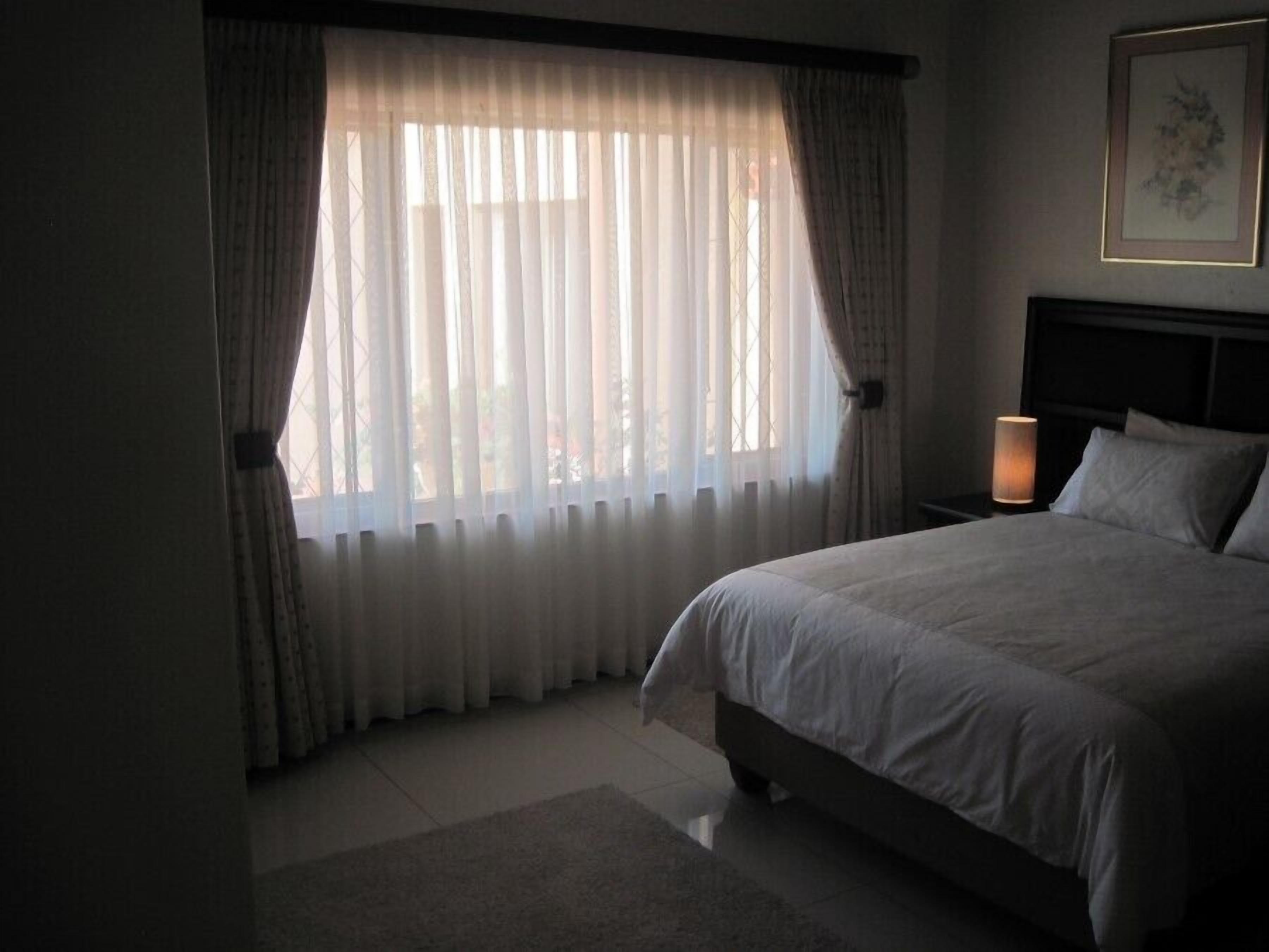 Quarto com cama grande, mesa de cabeceira com abajur, cortinas leves e um quadro na parede.