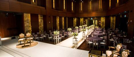 Uma ampla sala de banquetes com uma longa mesa posta para um evento formal, rodeada por cadeiras elegantes e lustres.