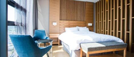 Quarto de hotel moderno com uma cama grande, uma poltrona azul, uma cabeceira de madeira e vista através de uma janela ampla.