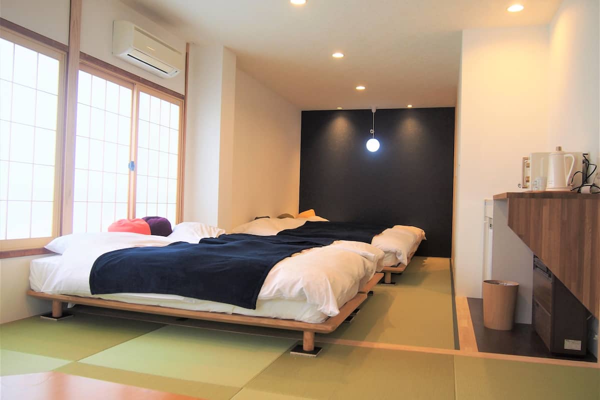 Quarto de hotel com cama, uma mesinha, lixeira e um espaço com cozinha compacta.