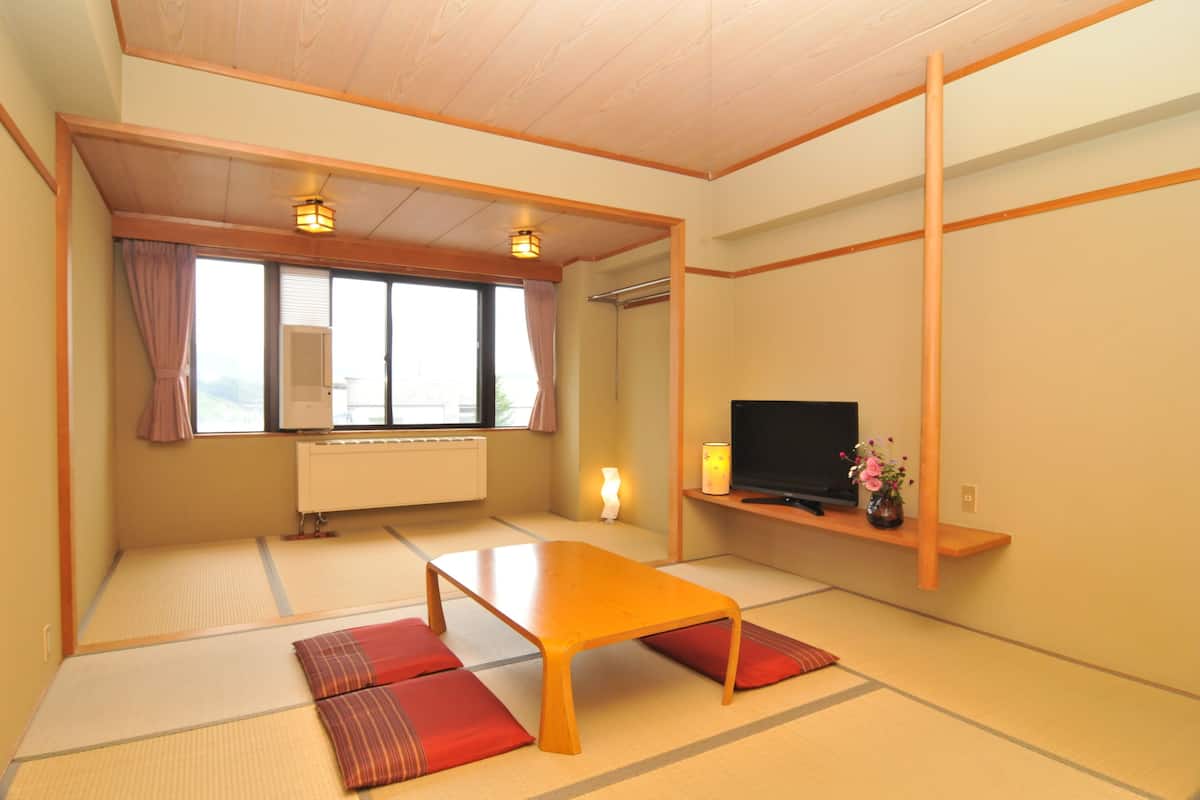 Um quarto japonês tradicional com tatames, uma mesa de madeira baixa, uma televisão em uma estante e uma janela com cortinas.