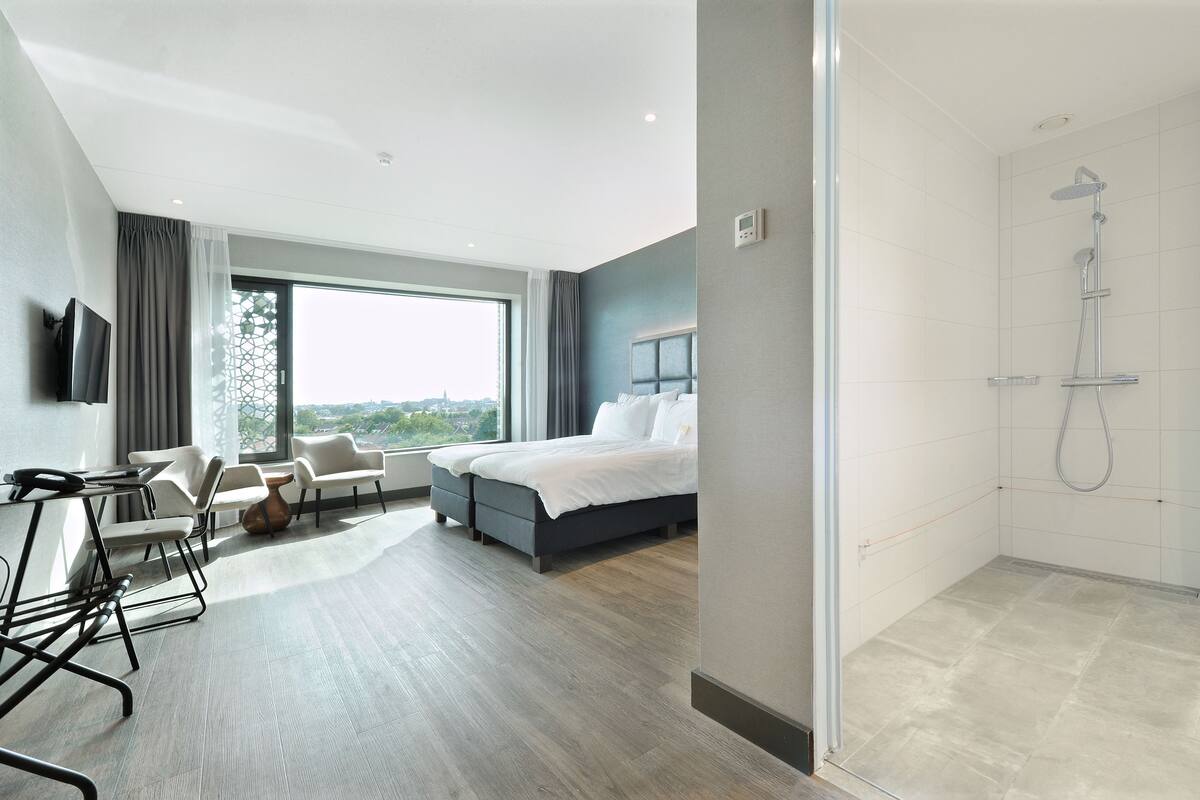 Quarto de hotel moderno com uma cama grande, uma mesa com cadeiras, uma TV e uma área de chuveiro.