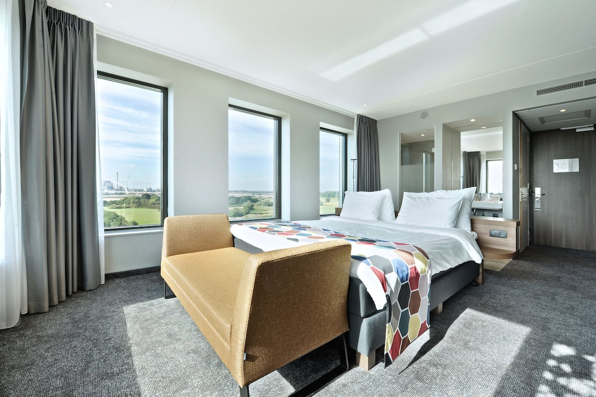 Quarto de hotel moderno com uma cama grande, uma cadeira, uma mesa e vista para o horizonte urbano.