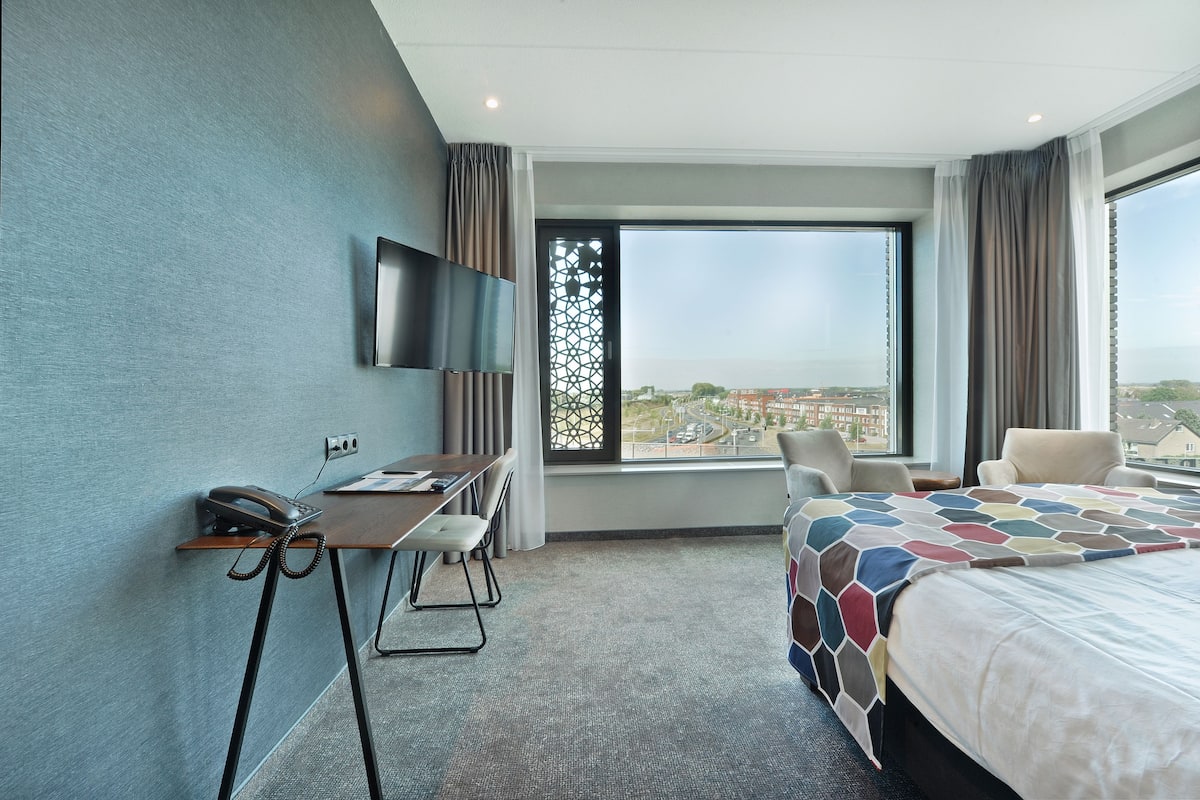 Quarto de hotel com uma cama grande, uma escrivaninha com telefone, uma TV fixada na parede e vista da cidade por uma janela ampla.