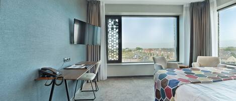 Quarto de hotel com uma cama grande, uma escrivaninha com telefone, uma TV fixada na parede e vista da cidade por uma janela ampla.