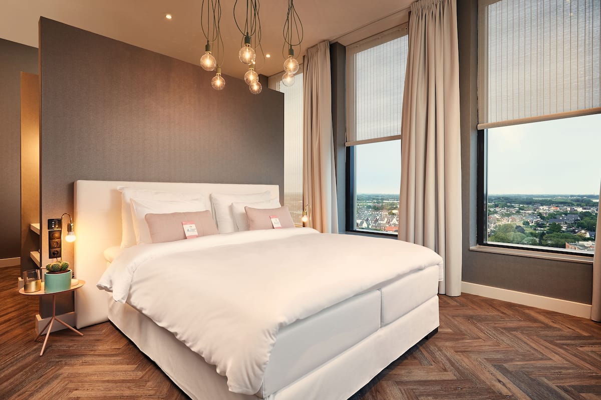 Quarto de hotel moderno com uma cama grande, uma mesa de cabeceira com abajur, vista da cidade através de amplas janelas e paisagem urbana visível lá fora.