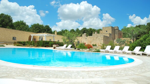 Piscina com espreguiçadeiras, um edifício de pedra e árvores ao fundo.