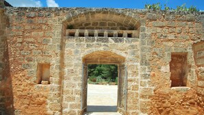 Uma entrada de pedra em arco com portão de metal, dando acesso a um pátio com piso de pedra e vegetação ao fundo.