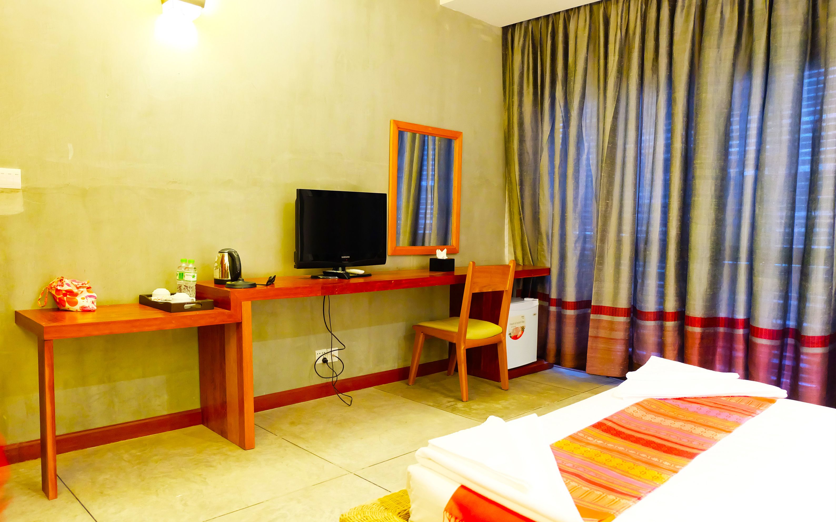 Quarto de hotel com cama, escrivaninha com televisão, cadeira e janela com cortinas.