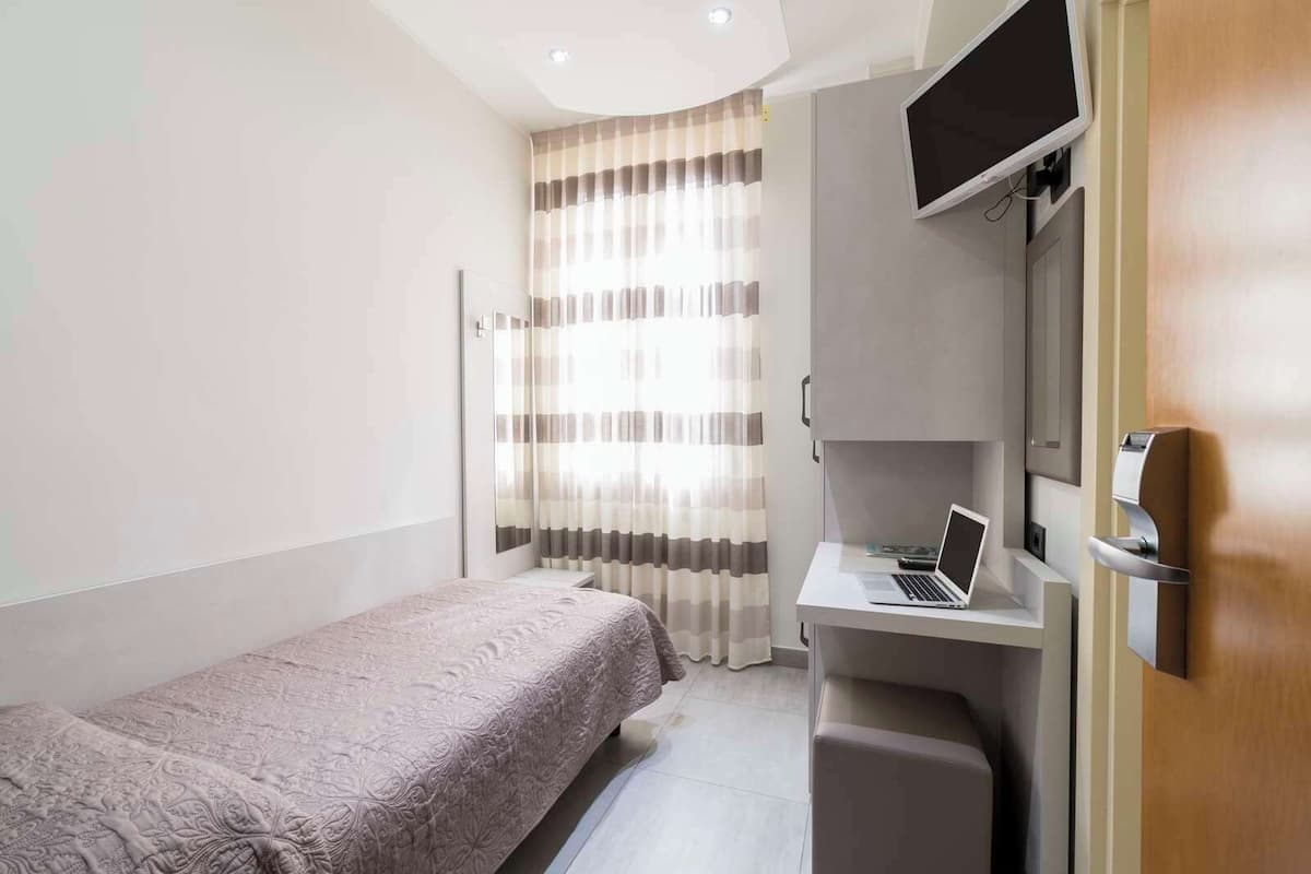 Um quarto pequeno e bem organizado, com uma cama de casal, uma escrivaninha com um laptop e uma televisão na parede.