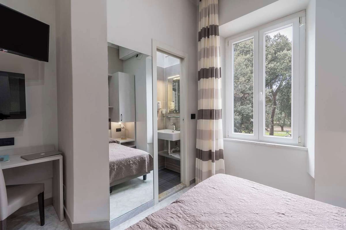 Quarto com cama, escrivaninha, televisão e janela com vista para árvores.