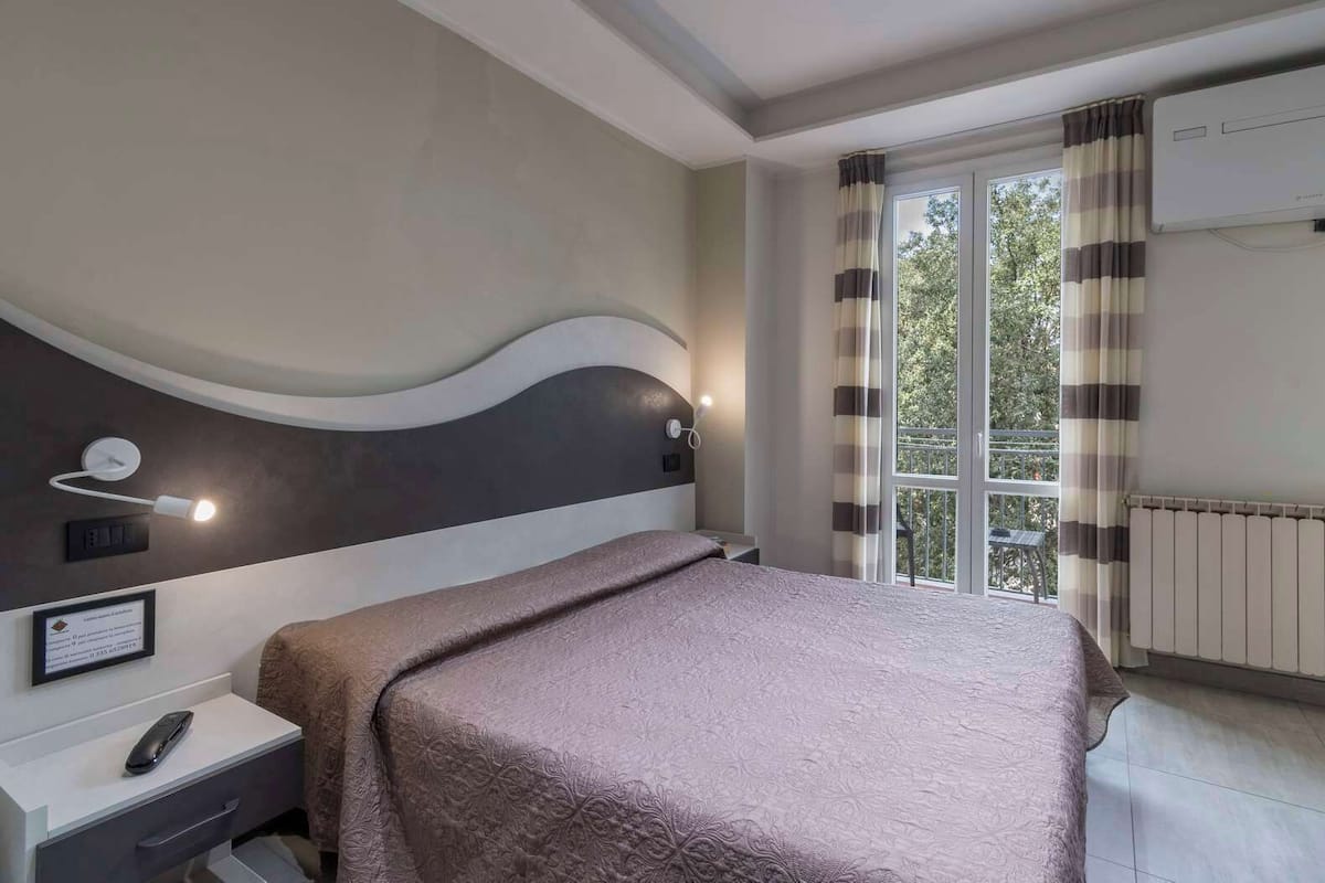 Quarto com cama, mesa de cabeceira, abajur e vista para o exterior pela janela.
