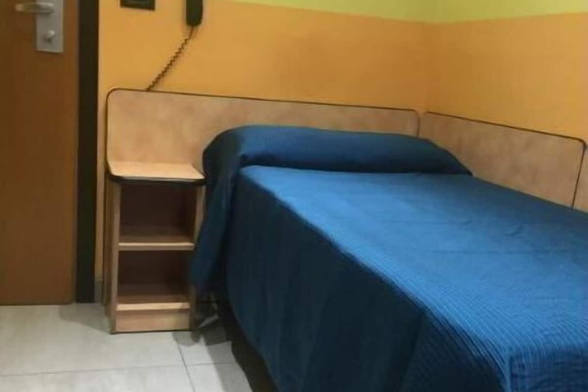 Quarto pequeno com uma cama de solteiro, cabeceira de madeira, colcha azul, mesa de cabeceira de madeira e telefone fixado na parede.
