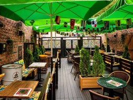 Um café ao ar livre com paredes de tijolo, mesas e cadeiras de madeira e guarda-sóis verdes.