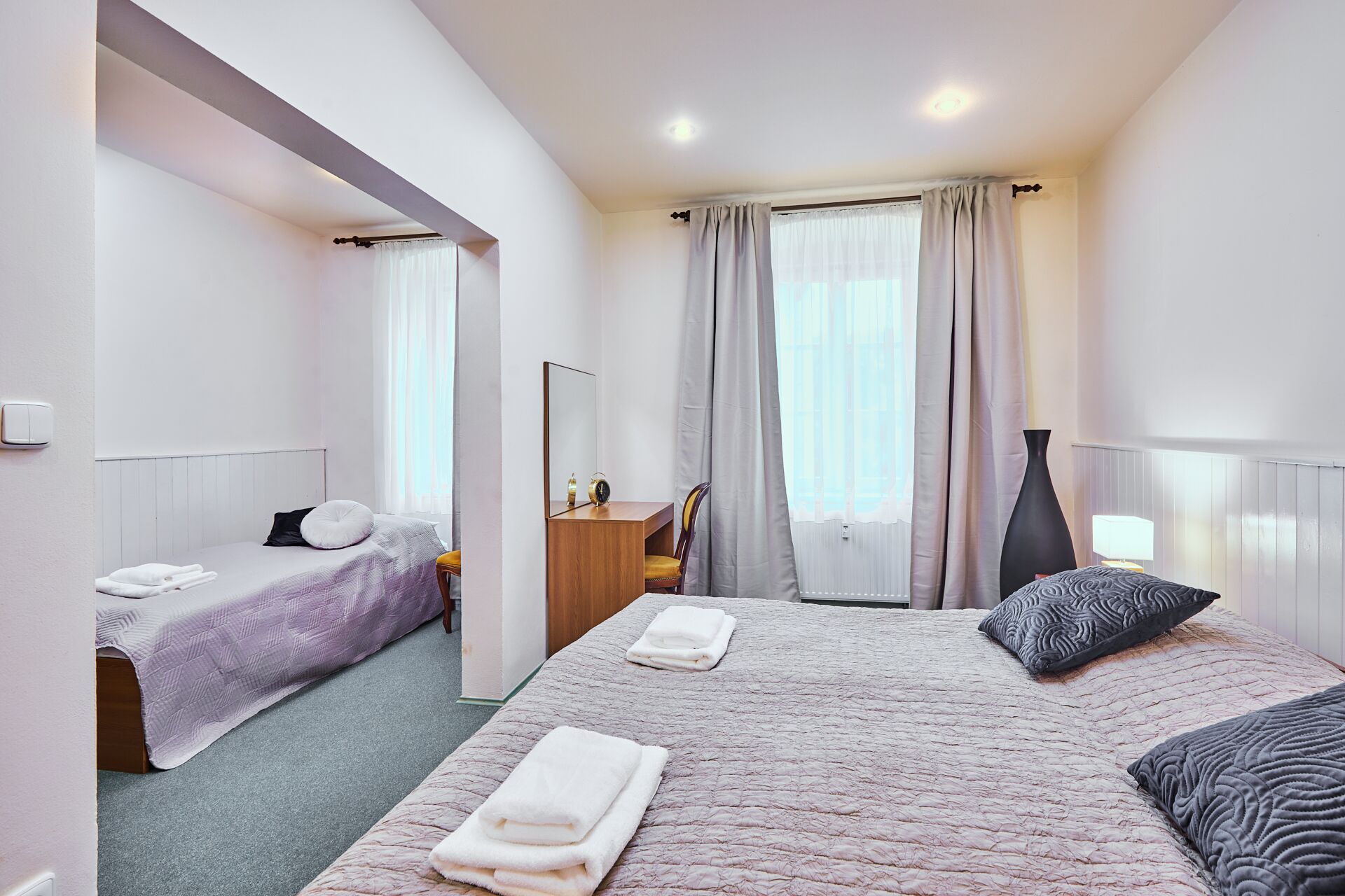 Quarto de hotel com duas camas, cômoda de madeira, espelho, luminária e janela com cortinas.