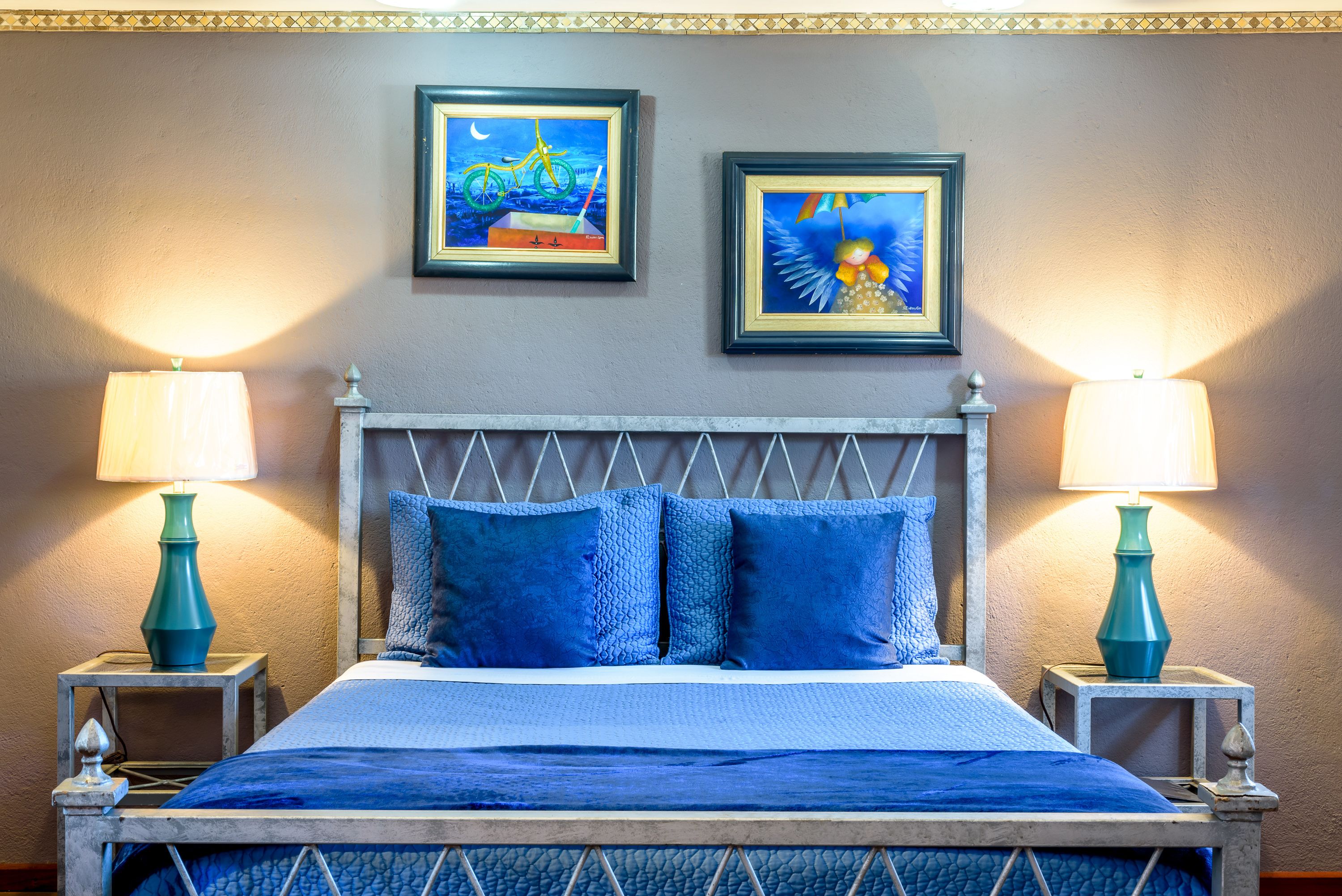 Uma cama com cabeceira azul e almofadas a combinar, com duas mesas de cabeceira e luminárias de cada lado.