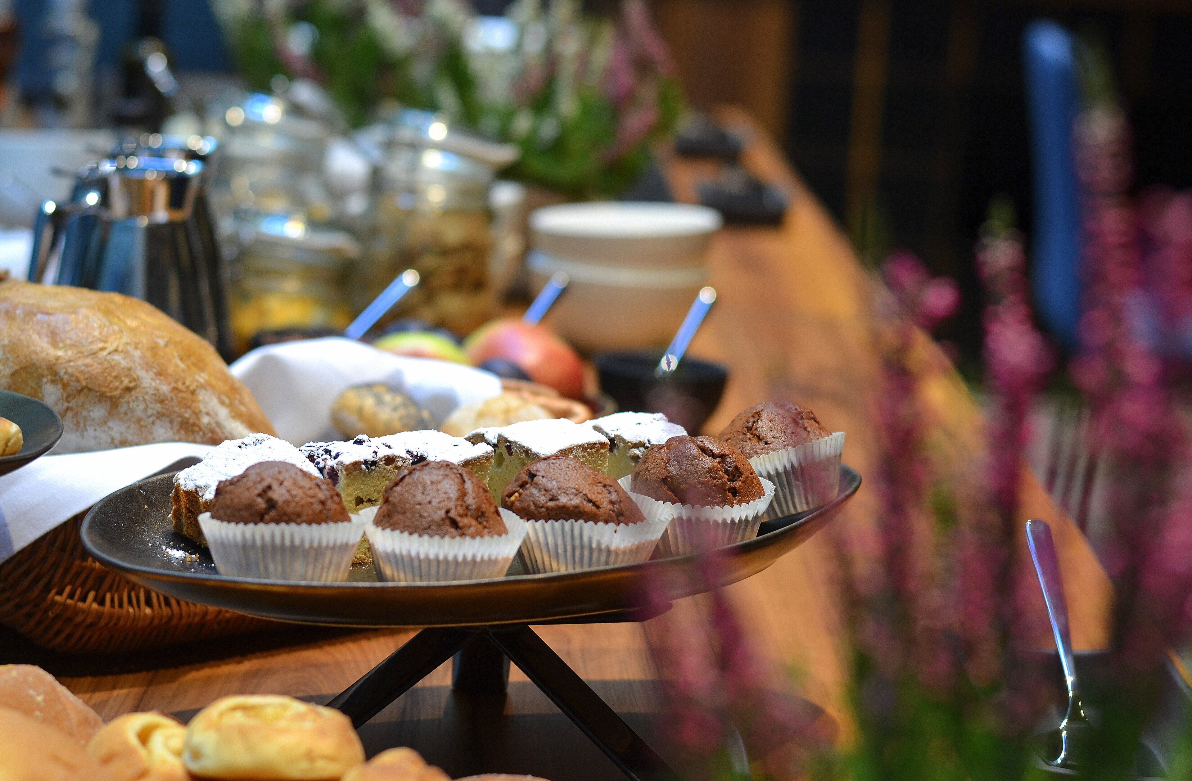 Uma bandeja com muffins e doces sobre uma mesa, com diversos alimentos e recipientes ao fundo.