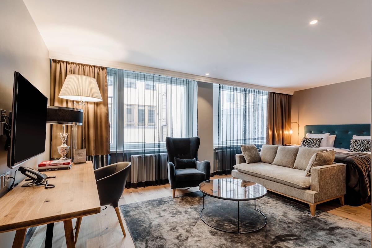 Quarto de hotel moderno com escrivaninha de madeira, poltrona preta, sofá bege e mesa de centro redonda.