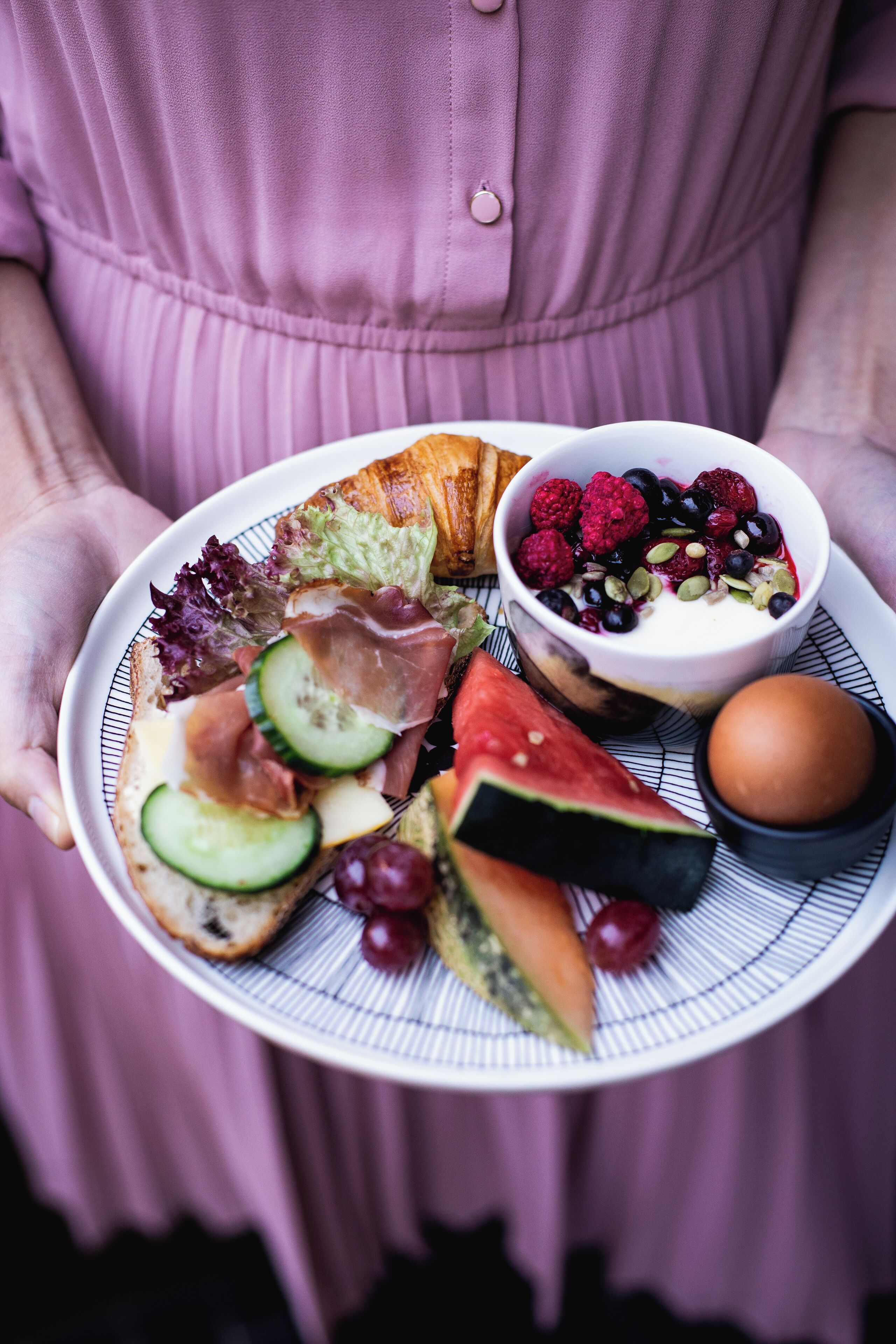 Um prato com um croissant, pepino fatiado, presunto, uvas, melancia e uma tigela de iogurte com frutas vermelhas e sementes.