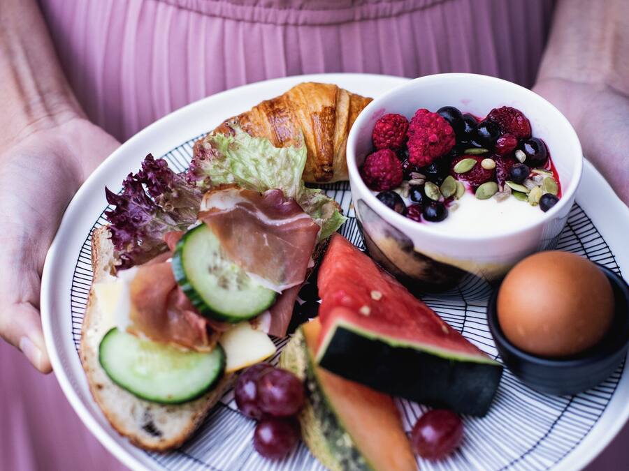 Um prato com um croissant, pepino fatiado, presunto, uvas, melancia e uma tigela de iogurte com frutas vermelhas e sementes.