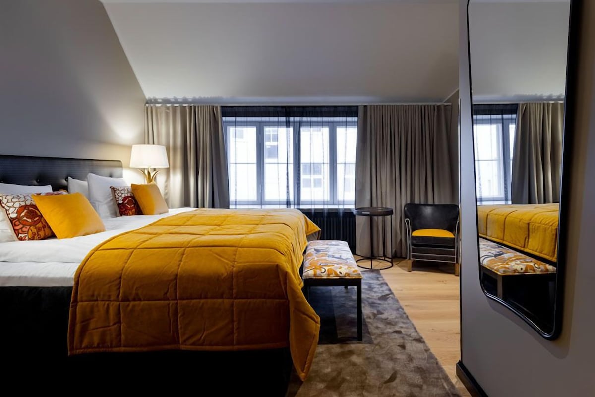 Quarto de hotel com uma cama grande, um banco, uma cadeira e uma mesa pequena.