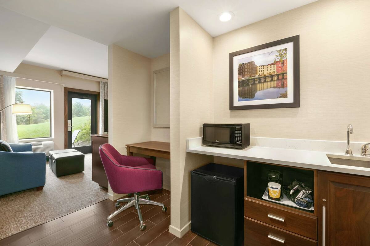 Quarto de hotel moderno com escrivaninha, cadeira, micro-ondas, frigorífico e uma imagem emoldurada da paisagem urbana na parede.