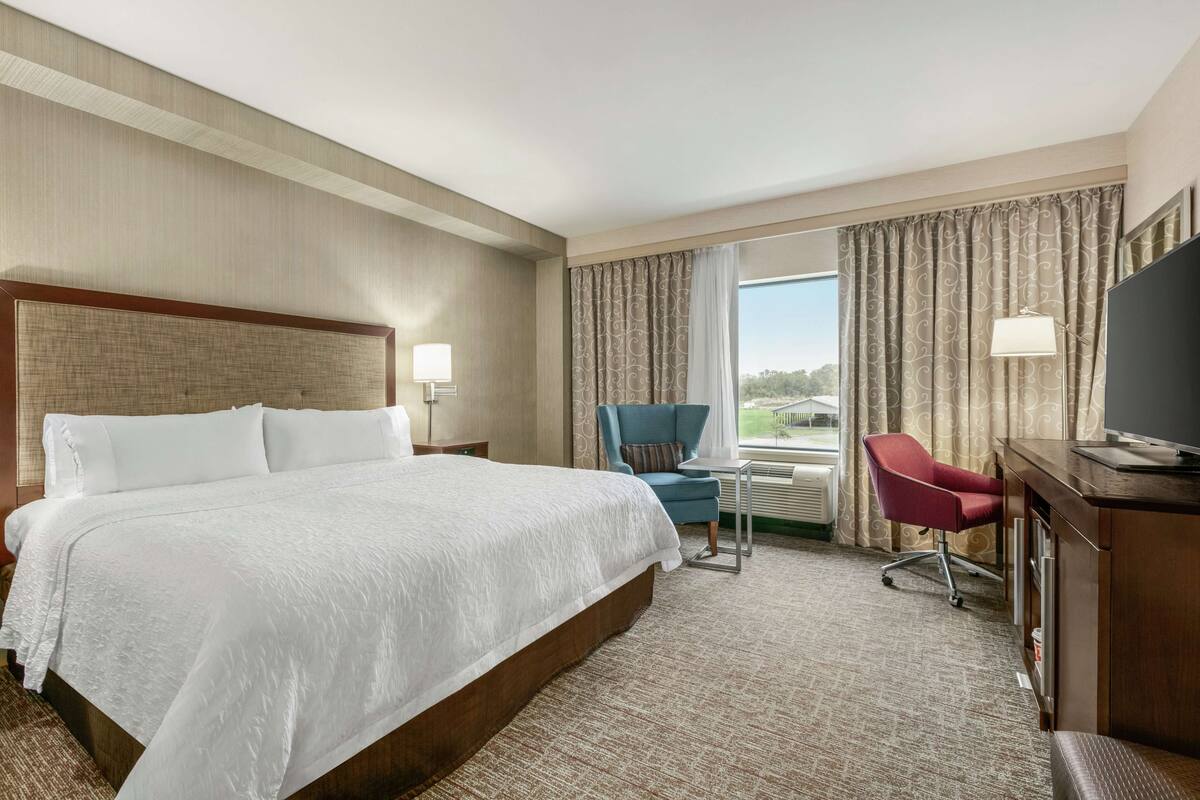 Quarto de hotel com uma cama grande, uma escrivaninha com cadeira, televisão e uma janela com cortinas.