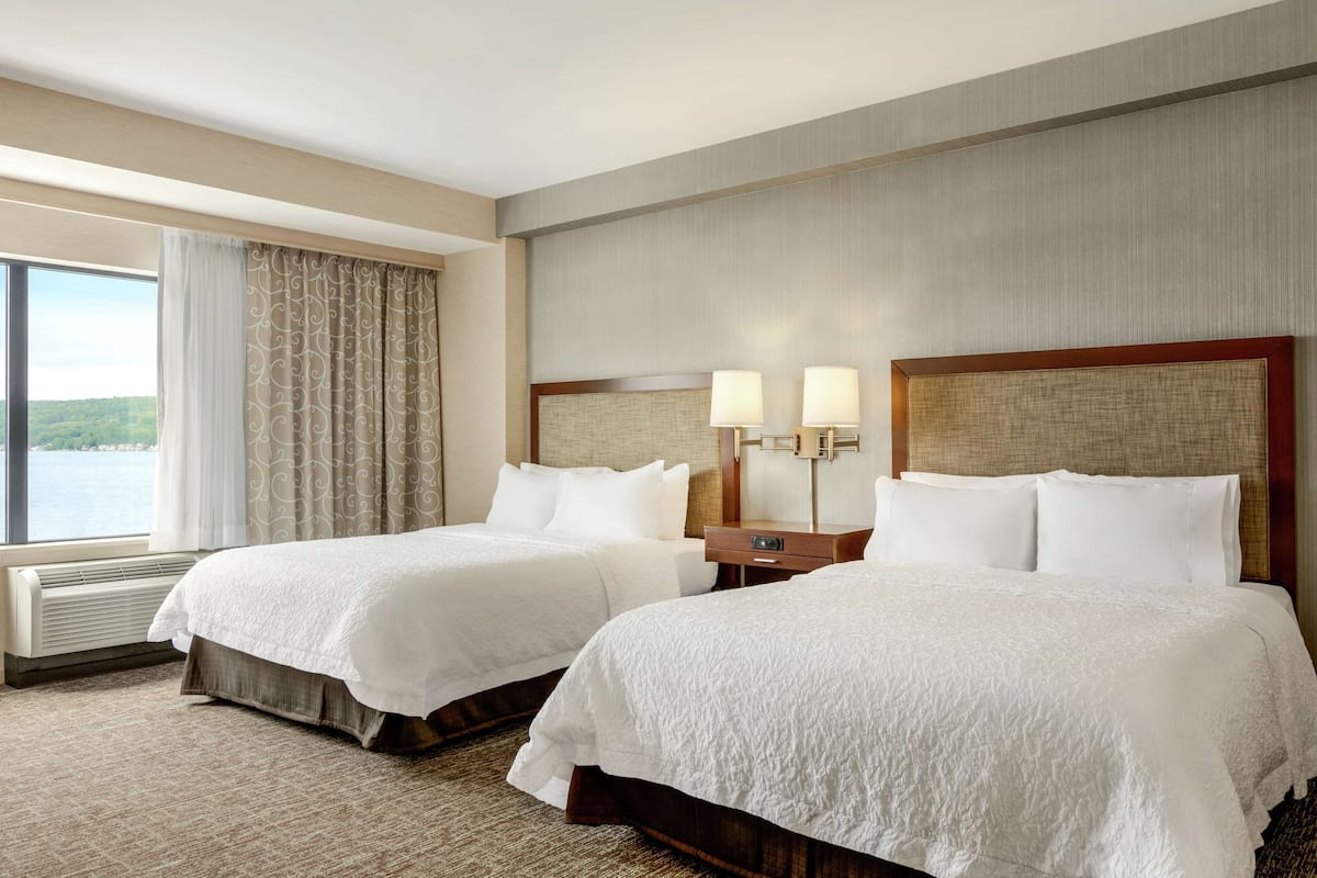 Quarto de hotel com duas camas, uma janela grande com cortinas e vista para o exterior, e uma luminária fixada na parede.