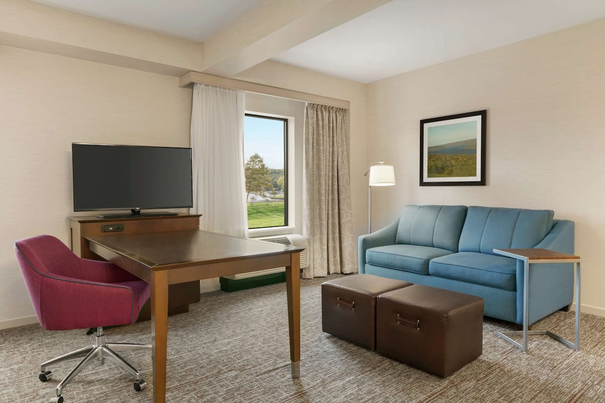Quarto de hotel com televisão, um sofá azul, uma cadeira vermelha, uma mesa de madeira e uma mesinha de cabeceira.
