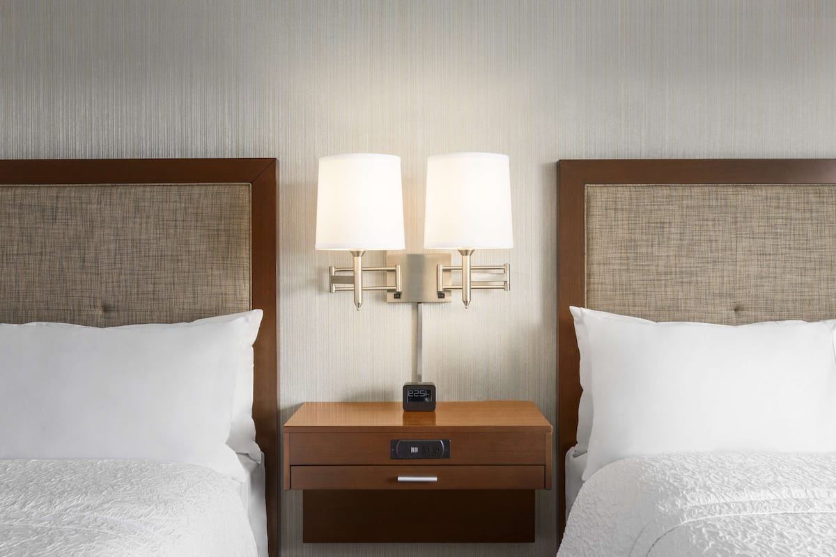 Quarto de hotel com duas camas, uma mesa de cabeceira com relógio digital e uma luminária na parede entre as camas.