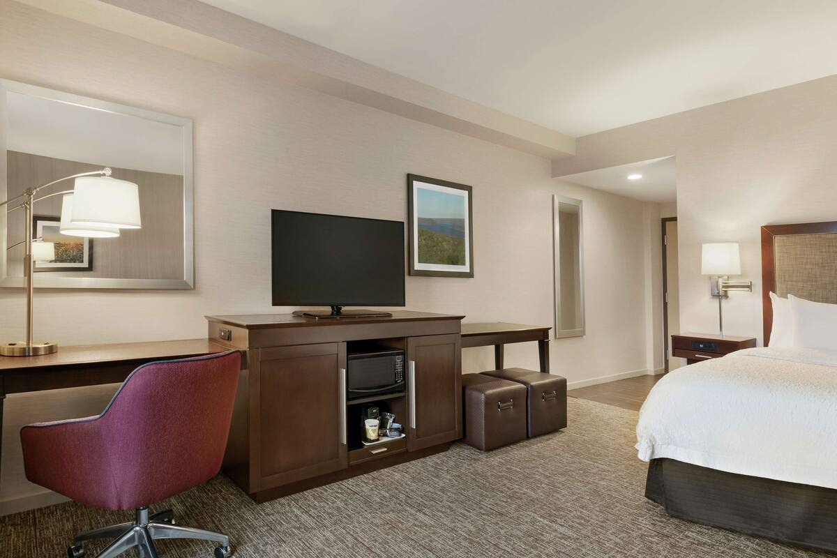 Quarto de hotel com cama, escrivaninha com computador, cadeira, televisão e um pequeno armário.