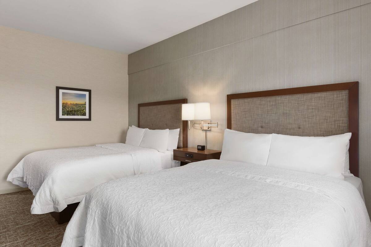 Quarto de hotel com duas camas, uma mesa de cabeceira com abajur e um quadro na parede.