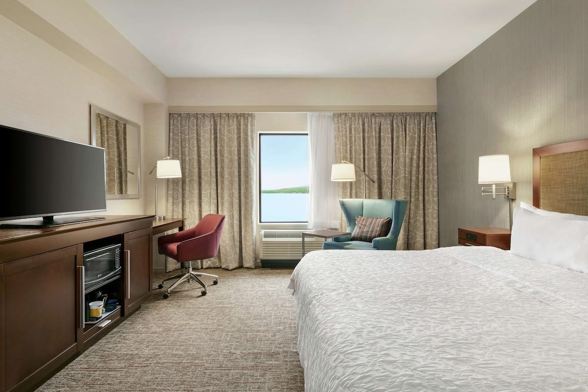 Quarto de hotel com uma cama grande, uma escrivaninha com televisão de tela plana, uma cadeira de escritório vermelha, um poltrona azul e vista para o exterior pela janela.