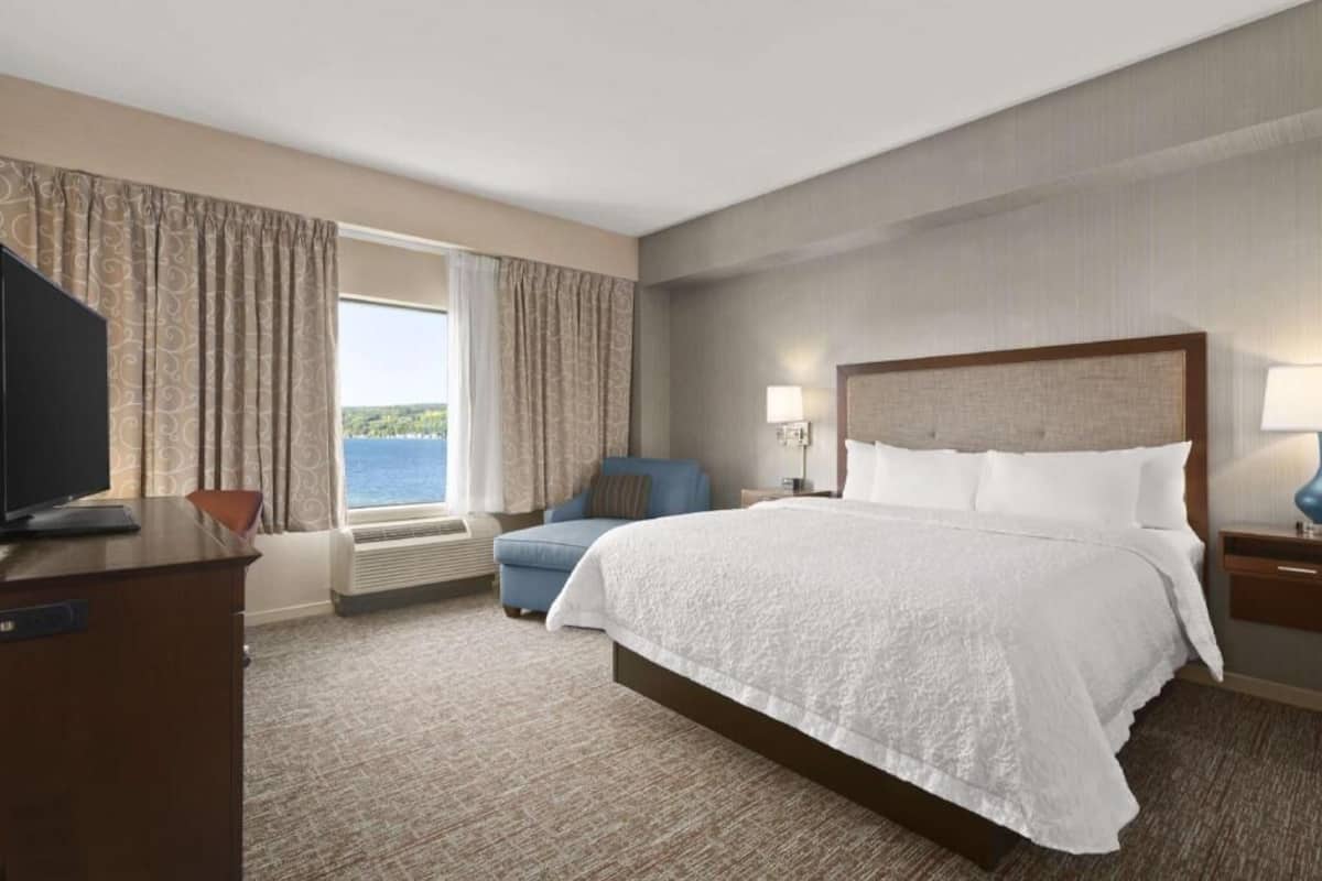 Quarto de hotel com uma cama grande, escrivaninha, cadeira, televisão e janela com cortinas.