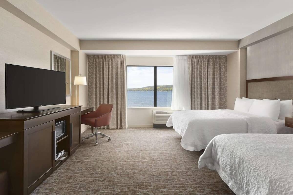 Quarto de hotel com televisão, duas camas, escrivaninha, cadeira e vista para a água.