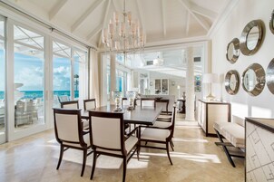 Uma ampla sala de jantar com uma mesa grande e cadeiras, um lustre e vista para o oceano através de grandes janelas.