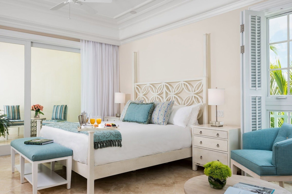 Quarto de hotel com cama, mesas de cabeceira, uma cadeira, uma pequena mesa com uma planta e uma janela com persianas.