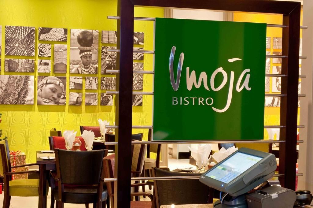 Interior de restaurante com um letreiro verde escrito 'Vmoja BISTRO', mesas com guardanapos brancos e um ecrã digital exibindo o menu.