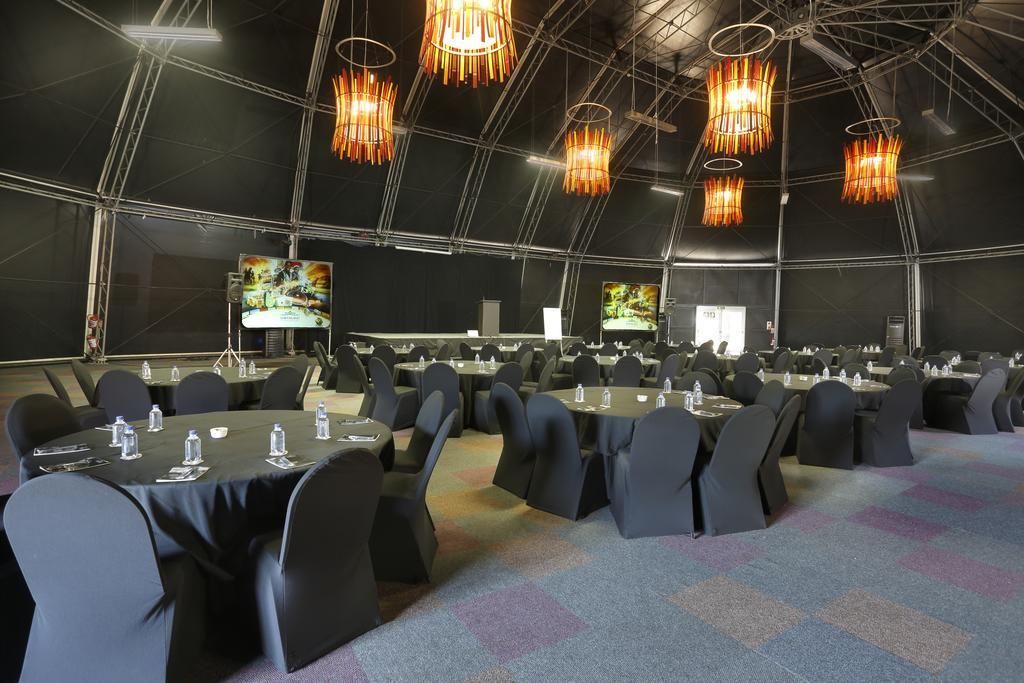 Sala de conferência com várias mesas e cadeiras, equipada com garrafas de água e placas de identificação.