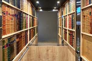 Um corredor estreito e bem iluminado, com prateleiras de madeira repletas de livros em ambos os lados.