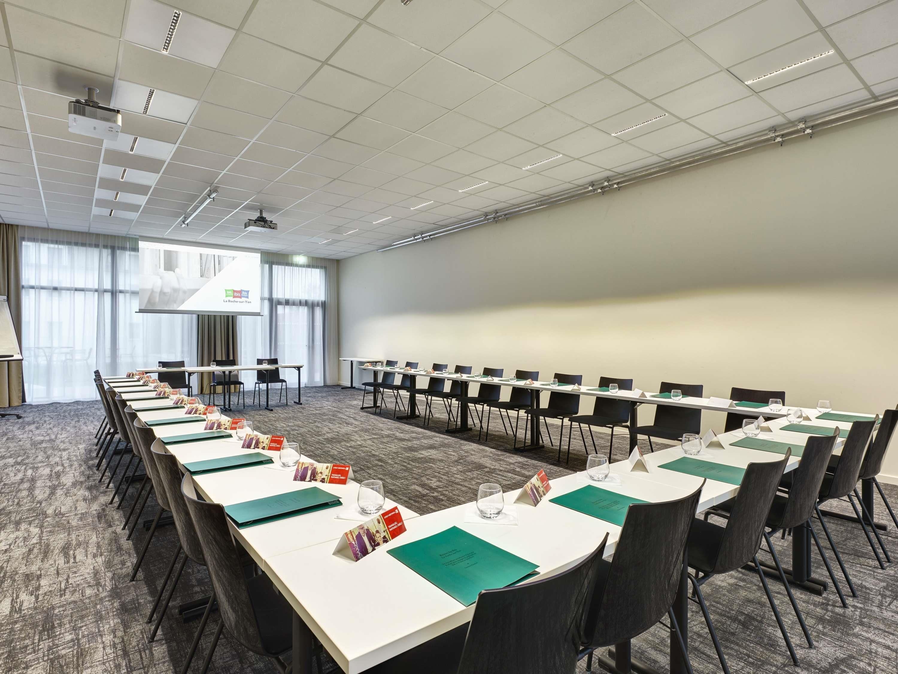 Uma sala de conferências com mesas compridas, cadeiras, um projetor e uma tela para apresentação.