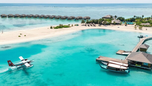 Vista aérea de um resort com bangalôs sobre a água, uma praia de areia e águas azul-turquesa cristalinas.