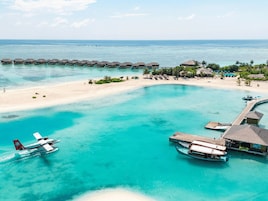 Vista aérea de um resort com bangalôs sobre a água, uma praia de areia e águas azul-turquesa cristalinas.