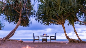 Cenário de praia com mesa e cadeiras sob palmeiras, com vista para o oceano.