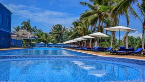 Piscina com azulejos de mosaico azul, rodeada por espreguiçadeiras e guarda-sóis, com palmeiras e céu claro ao fundo.