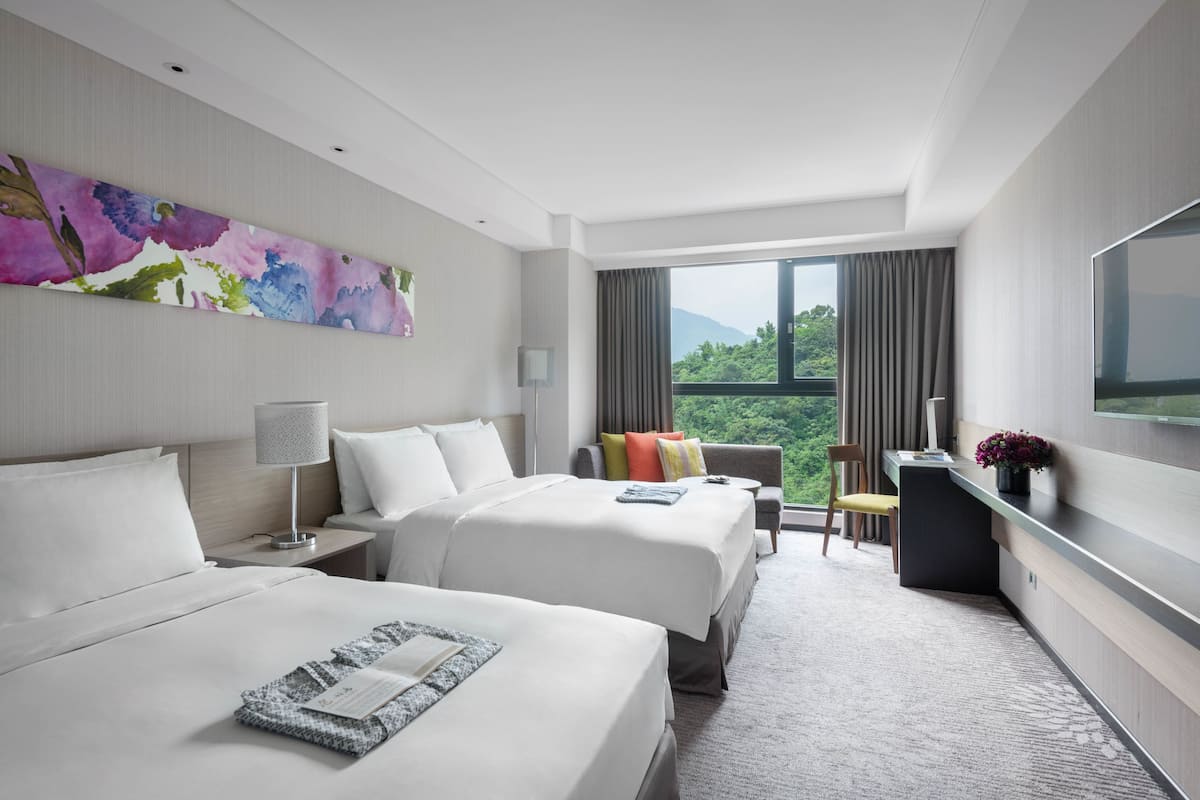 Quarto de hotel com duas camas, uma escrivaninha, uma cadeira, uma TV e uma janela grande com vista para vegetação.